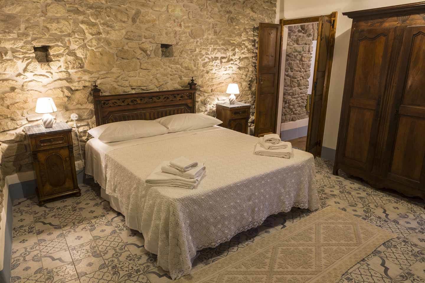 Uma cama com colcha branca, duas luminárias de cabeceira, uma cabeceira de madeira e uma parede de pedra.