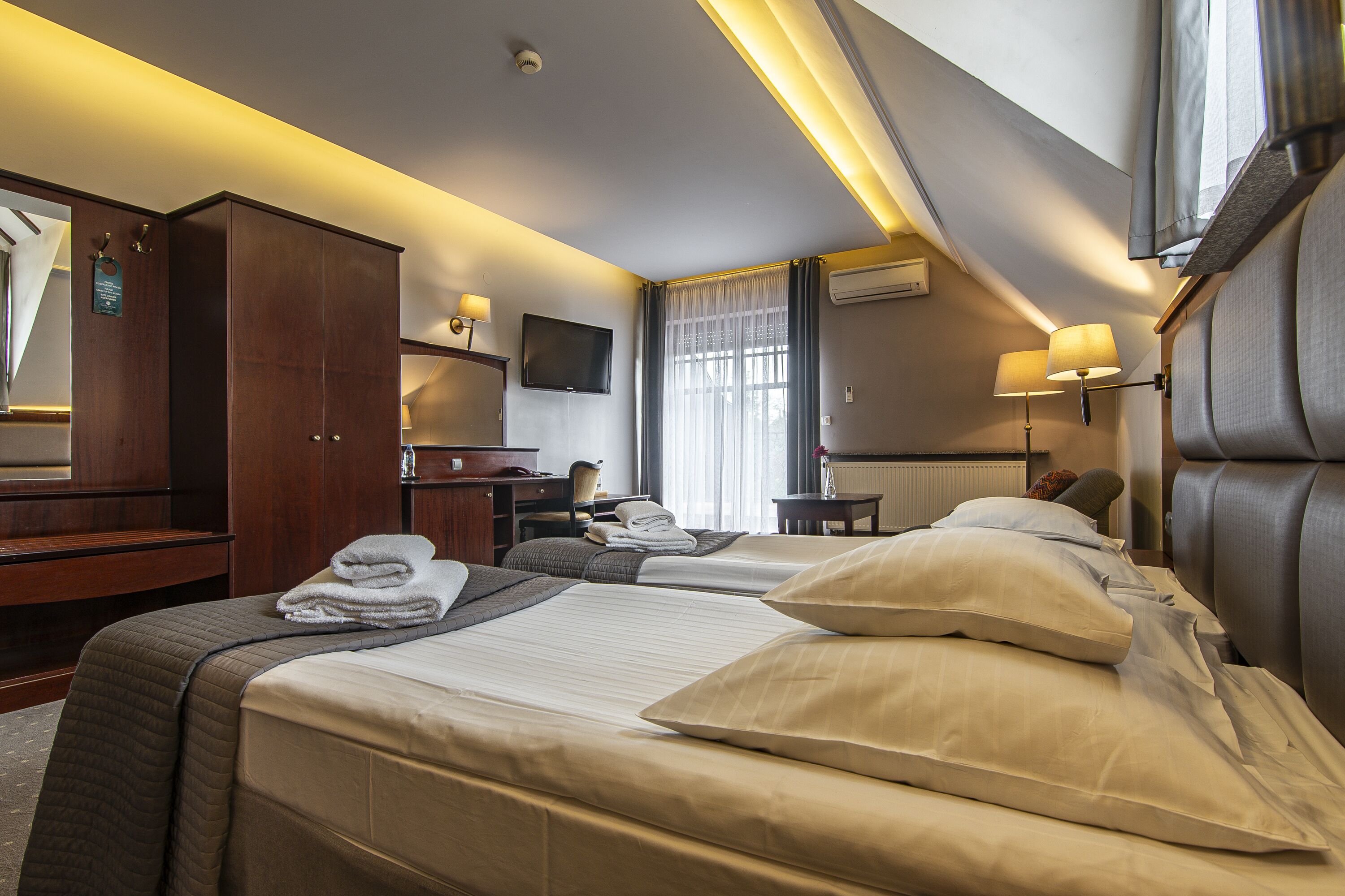 Quarto de hotel com duas camas, uma escrivaninha, uma cadeira, uma televisão e um guarda-roupa.
