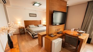 Quarto de hotel compacto com cama, TV fixada em painel de madeira, pequena cozinha e escrivaninha com cadeira.