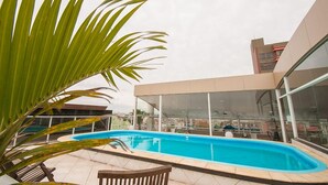 Piscina no terraço com estrutura de vidro, cercada por palmeiras e edifícios modernos.
