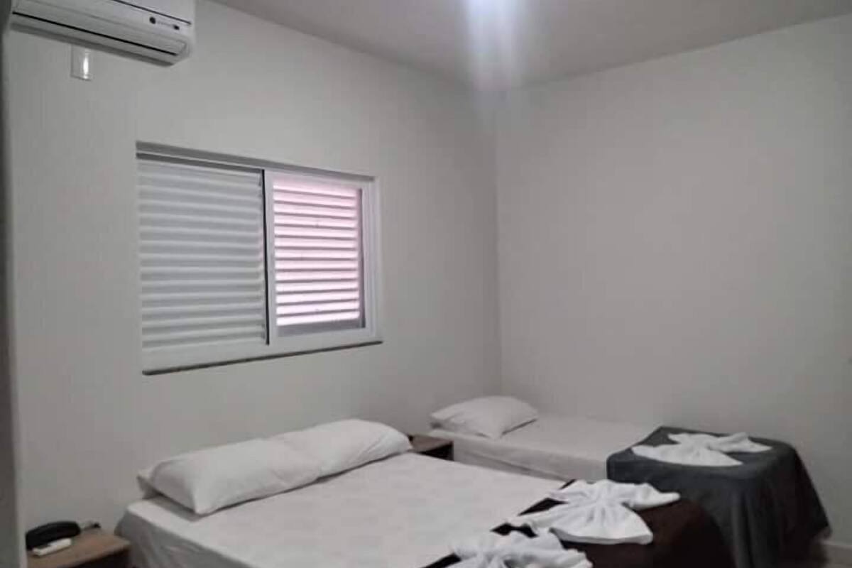 Quarto pequeno e limpo, com duas camas, uma mesa de cabeceira e janela com persianas.