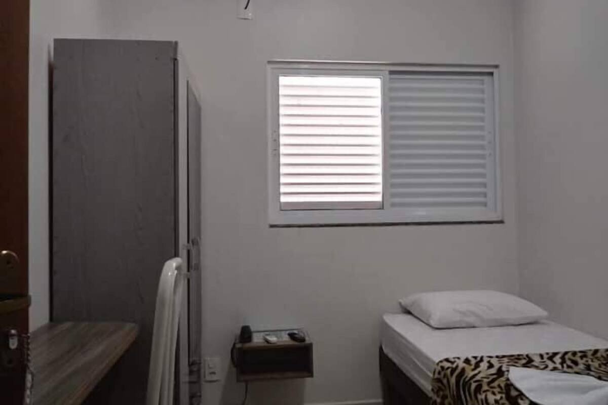 Quarto pequeno e simples com cama, janela com persianas, radiador e uma mesa pequena.