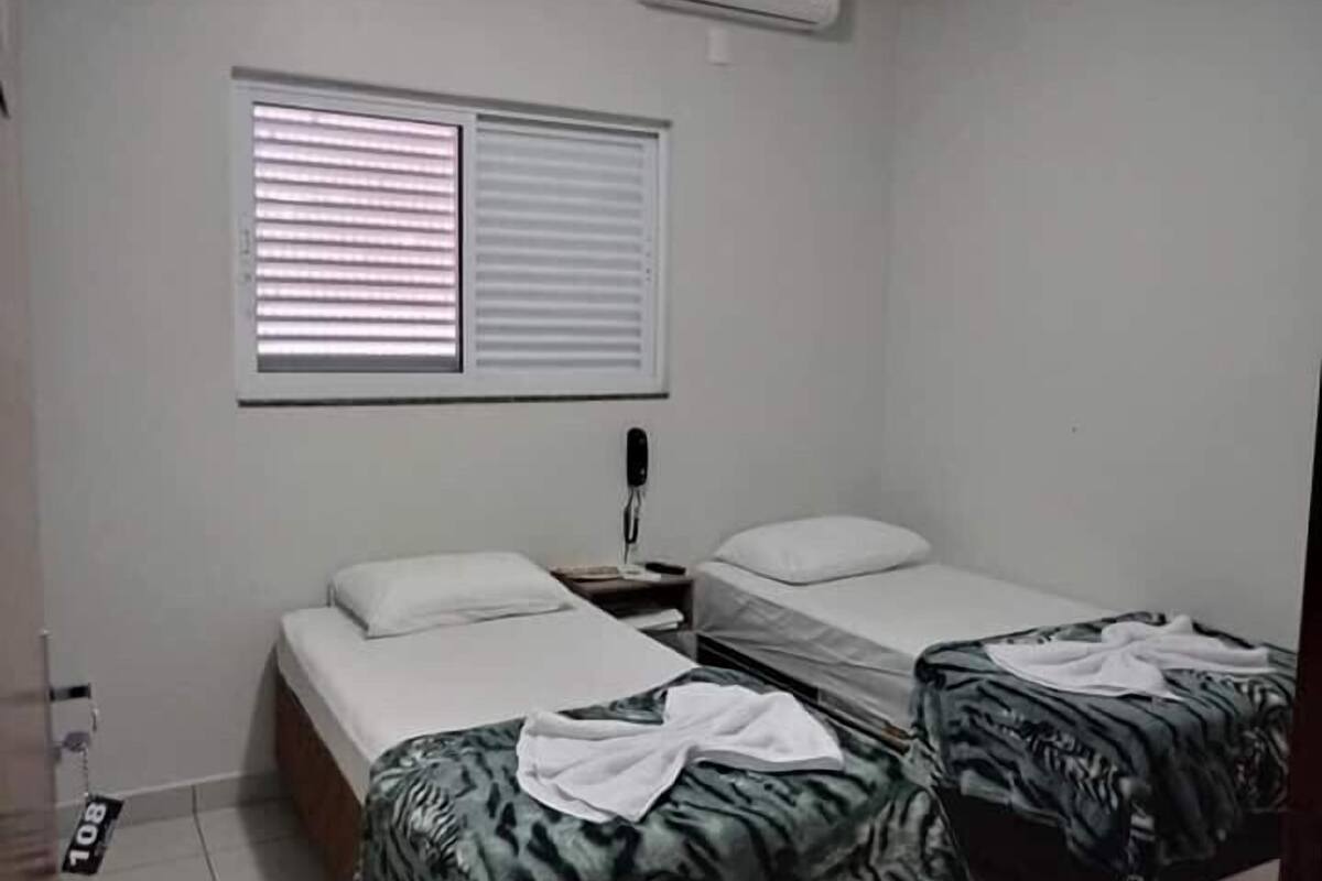 Quarto pequeno e limpo com duas camas, janela com persianas, mesa de cabeceira com telefone e porta com o símbolo de número.