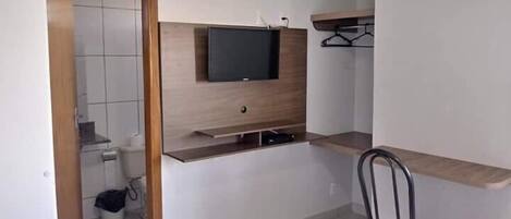 Quarto compacto com televisão integrada, um pequeno espaço para sentar e uma estante de madeira.