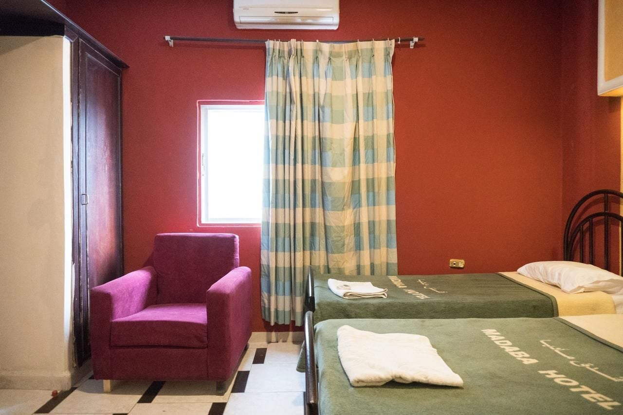 Quarto com duas camas, parede vermelha, janela com cortina e uma cadeira roxa.