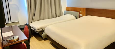 Quarto de hotel com duas camas de solteiro, uma escrivaninha com cadeira, um cabideiro e um quadro na parede.