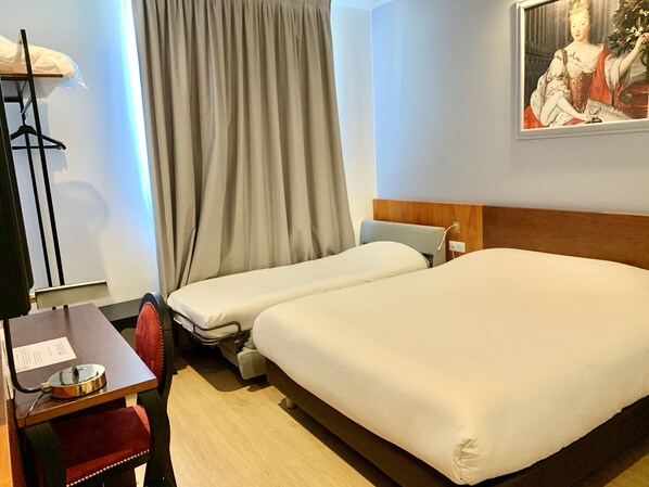 Quarto de hotel com duas camas de solteiro, uma escrivaninha com cadeira, um cabideiro e um quadro na parede.