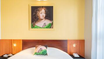Quarto de hotel com cama, mesa de cabeceira, um quadro na parede e almofada com motivos florais.