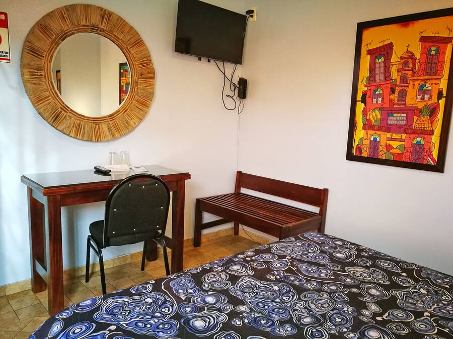 Quarto com escrivaninha de madeira, uma cadeira, cama com colcha estampada, espelho redondo, televisão fixada na parede e uma pintura colorida.
