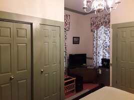 Quarto com duas portas verdes, uma televisão sobre um suporte, uma pequena mesa de madeira e cortinas florais.