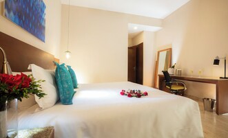 Quarto de hotel com cama, escrivaninha, espelho e um vaso com flores.