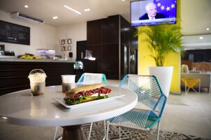 Um lobby moderno de hotel com uma mesa redonda, um sanduíche, uma bebida da Starbucks e uma tela de TV exibindo uma notícia.