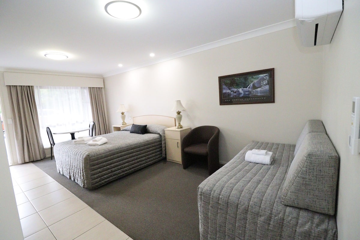 Quarto de hotel com duas camas, uma mesinha de cabeceira, uma cadeira, uma janela com cortinas e um quadro na parede.