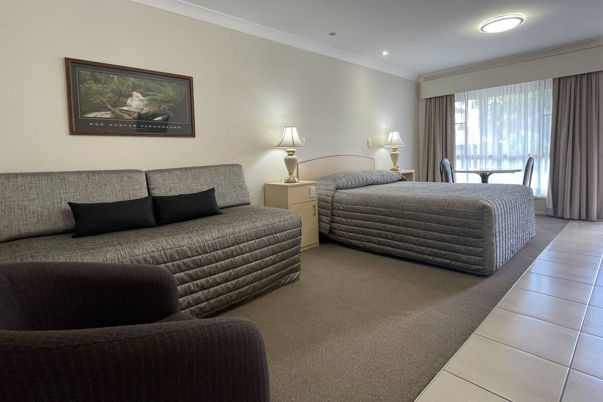 Quarto de hotel com sofá cinzento, um sofá menor, uma cama, mesa de cabeceira com luminárias e um quadro emoldurado na parede.