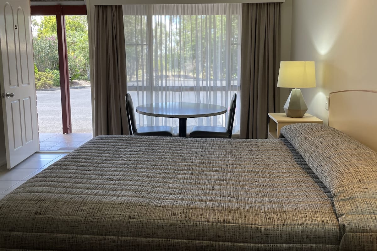 Quarto de hotel com cama, mesa, cadeiras, abajur e vista para o verde pela janela.