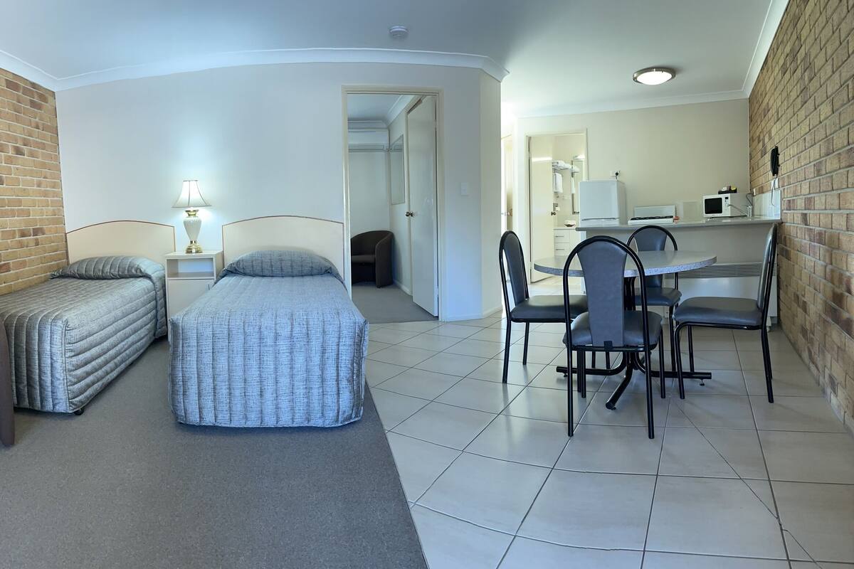 Quarto com duas camas de solteiro, uma escrivaninha com cadeira, uma TV fixada numa parede de tijolos e uma mesinha de cabeceira com um controle remoto.