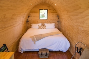 Acomodação acolhedora, em um único ambiente, com teto inclinado, cama com travesseiros e coberta, uma mesa pequena com cesta e janela com vista.