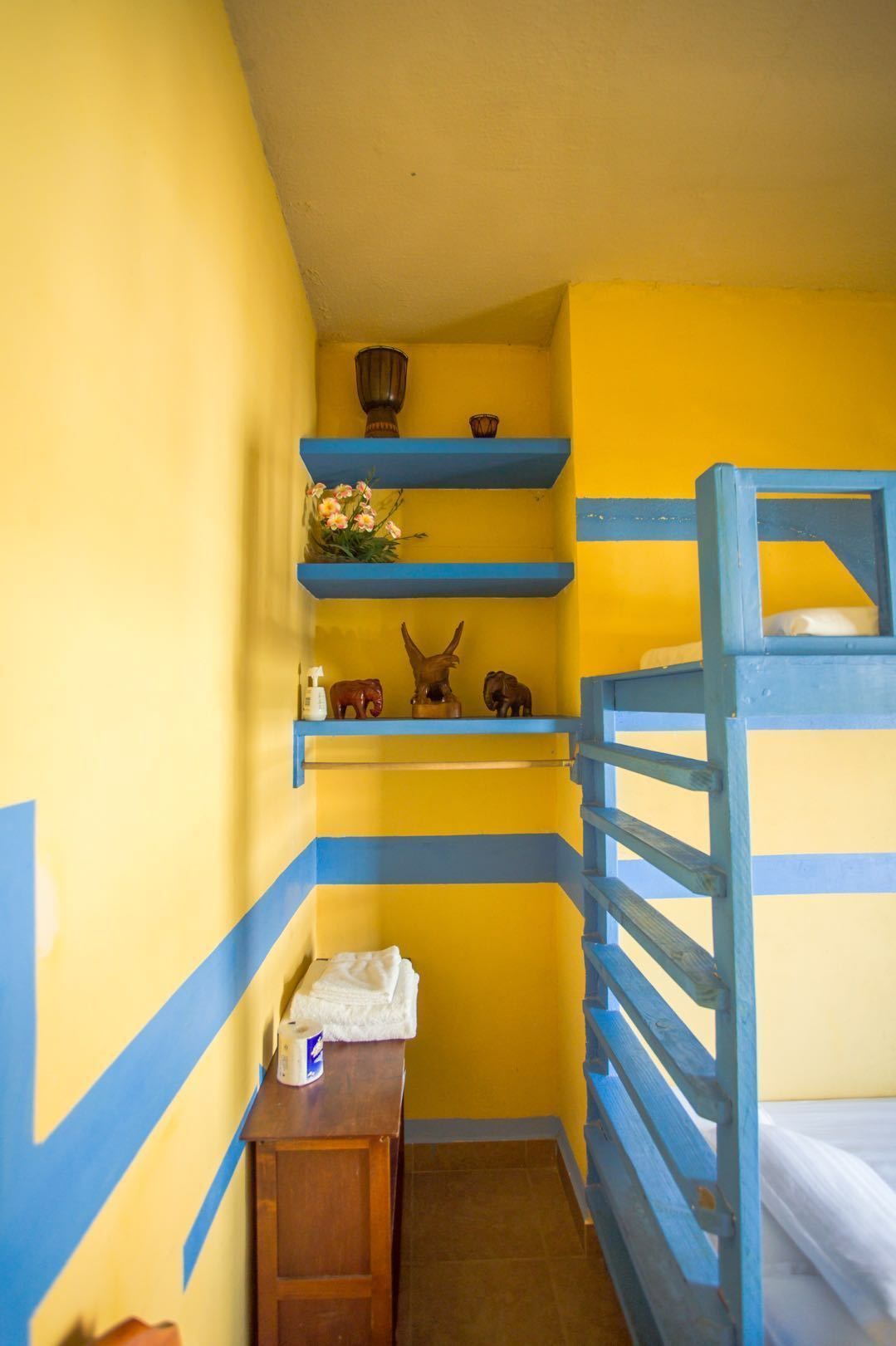 Um quarto pequeno e colorido com uma cama beliche, prateleiras com objetos decorativos e uma pequena mesa com uma garrafa.