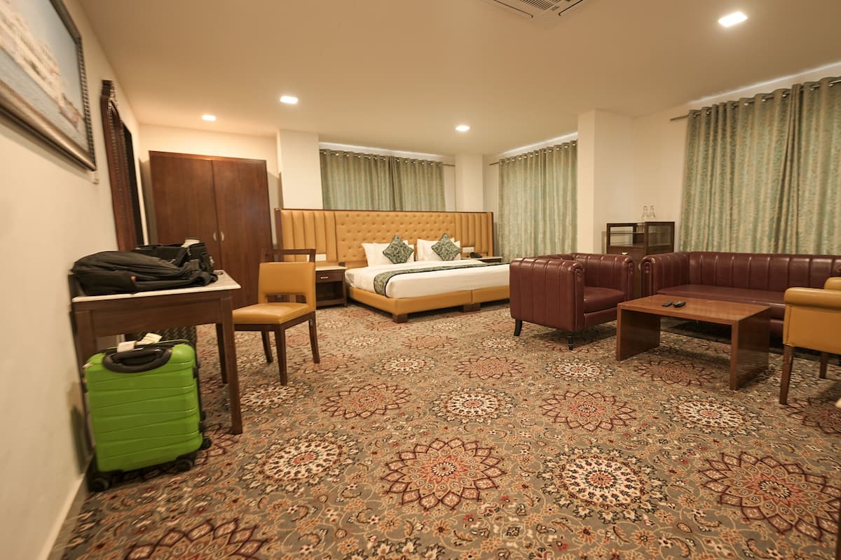 Quarto de hotel com uma cama, um sofá, uma cadeira, uma mesinha, uma escrivaninha e uma mala verde.