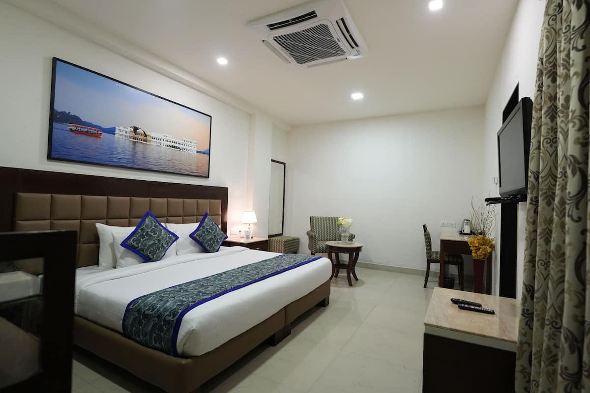 Quarto de hotel com cama, escrivaninha, cadeira, televisão e um quadro na parede.