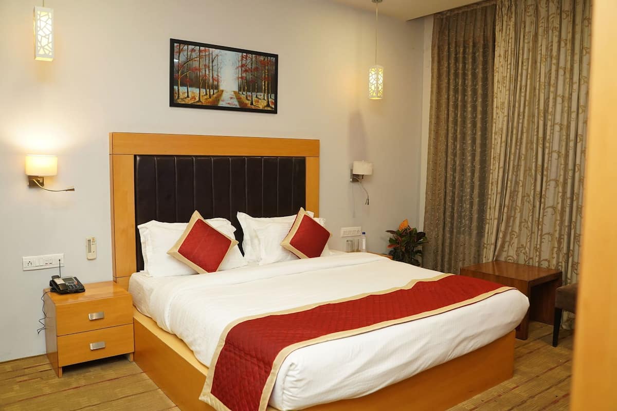 Quarto de hotel com uma cama grande, cabeceira, abajures, mesa de cabeceira, telefone, um quadro na parede e cortina.