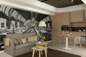 Uma sala de estar moderna com um sofá cinza, uma pequena mesa com um livro e uma cadeira de vidro transparente. Um grande mural em preto e branco de uma estação de trem está na parede.