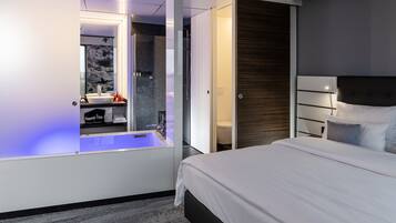 Quarto de hotel moderno com uma cama grande, chuveiro embutido e vista para o horizonte urbano através de uma parede de vidro.