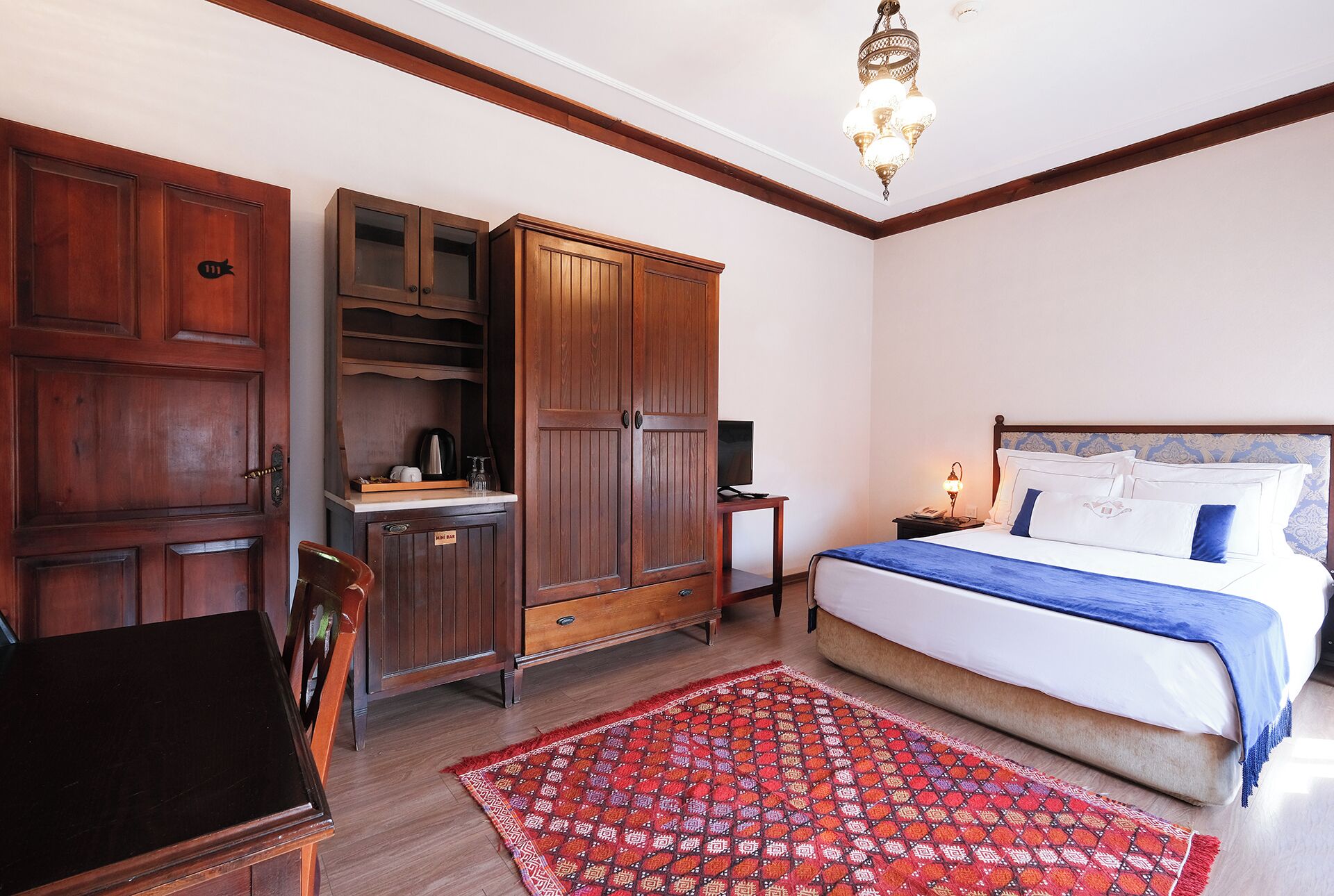 Quarto com cama, guarda-roupa de madeira, escrivaninha e tapete vermelho estampado.