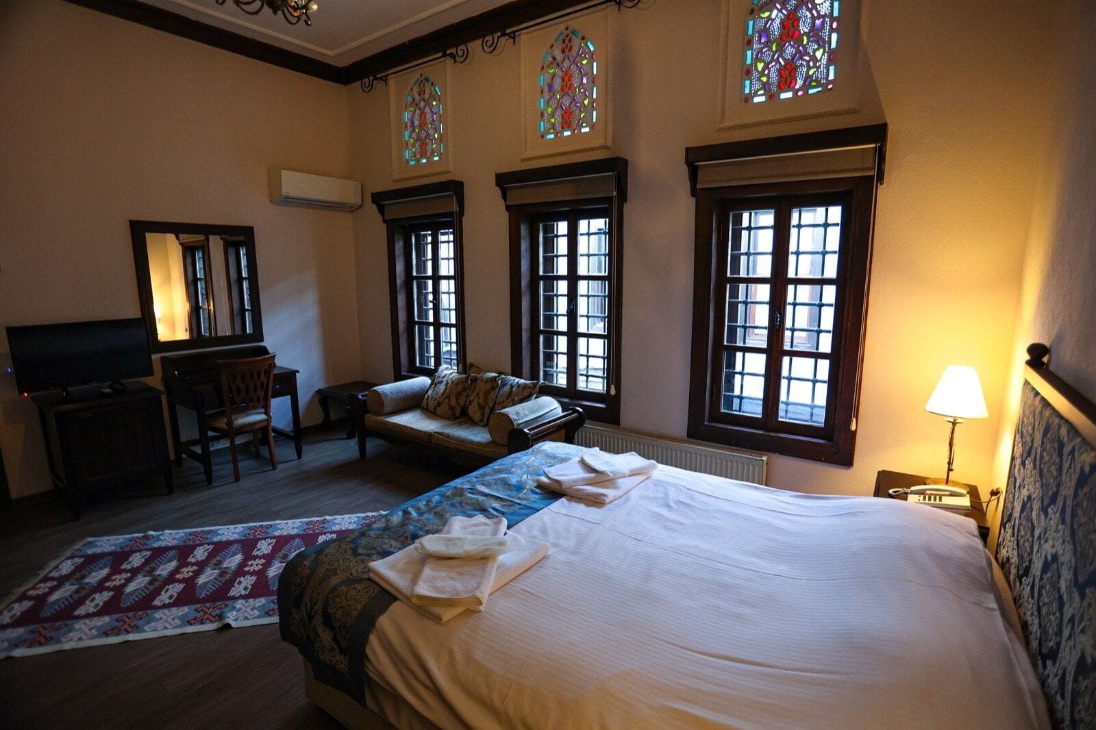 Quarto de hotel com uma cama grande, uma escrivaninha, uma cadeira e uma televisão. Há janelas de vitrais e um abajur na mesa de cabeceira.