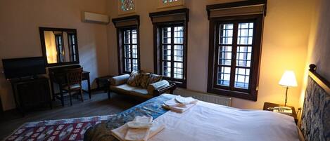 Quarto de hotel com uma cama grande, uma escrivaninha, uma cadeira e uma televisão. Há janelas de vitrais e um abajur na mesa de cabeceira.
