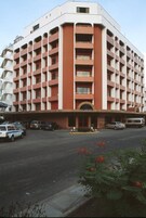 Um hotel de vários andares com entrada central e um estacionamento em frente.