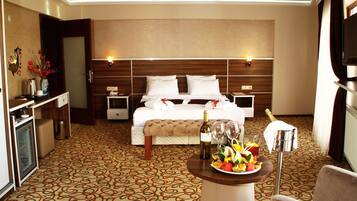 Quarto de hotel com cama, mesas de cabeceira, minibar, uma pequena mesa com uma tábua de frutas e vinho, e uma televisão.