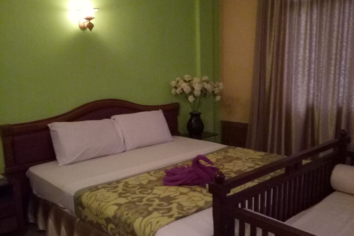 Quarto de hotel com cama, mesa de cabeceira, um vaso com flores e janela com cortinas.