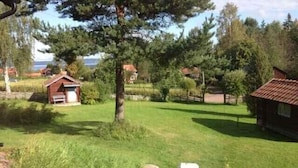 Paisagem rural com uma pequena cabana de madeira, um quintal cercado e uma grande árvore.