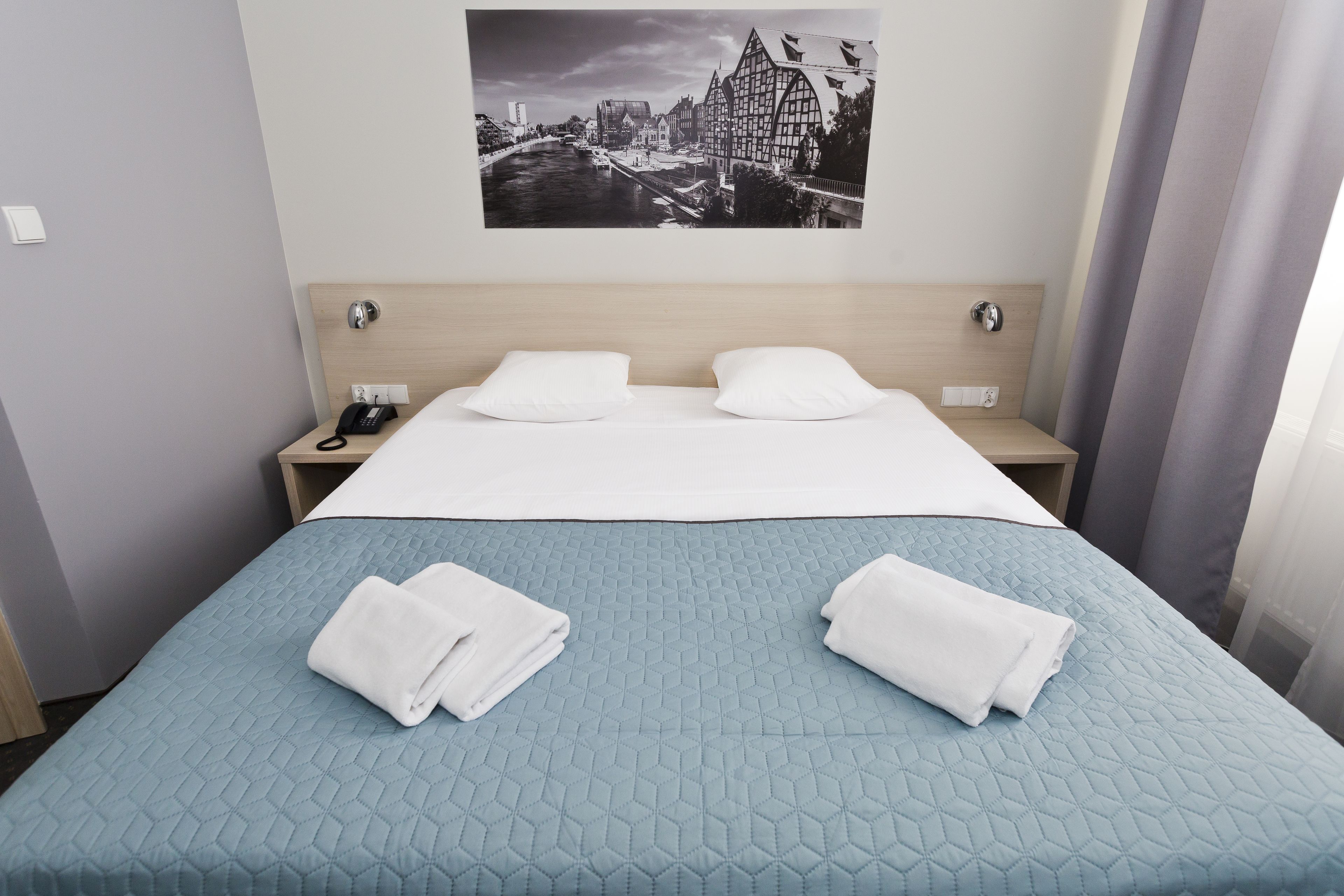 Quarto de hotel com uma cama grande, duas almofadas, mesa de cabeceira com telefone e uma fotografia de paisagem urbana na parede.