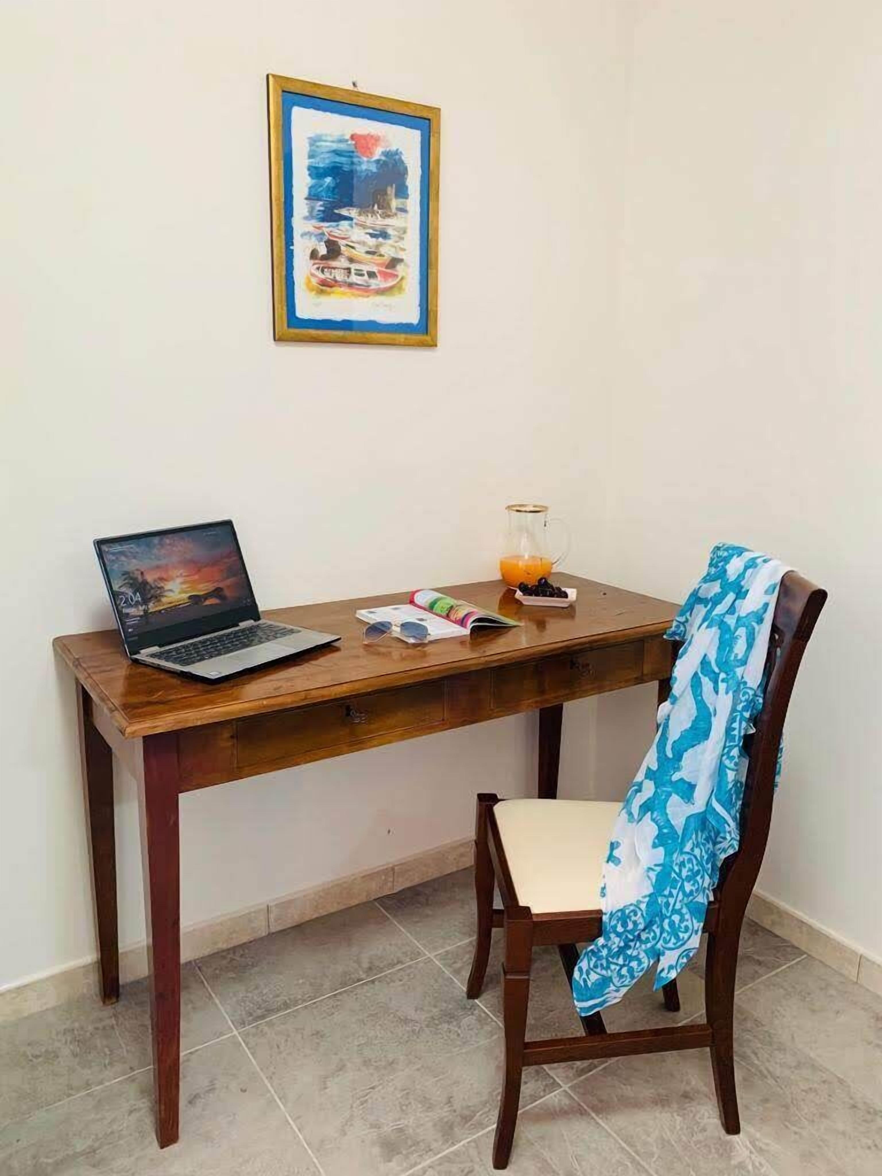 Uma mesa de madeira com um laptop, um jarro de vidro com sumo de laranja e um livro. Uma cadeira com um tecido estampado em azul e branco está posicionada em frente à mesa.