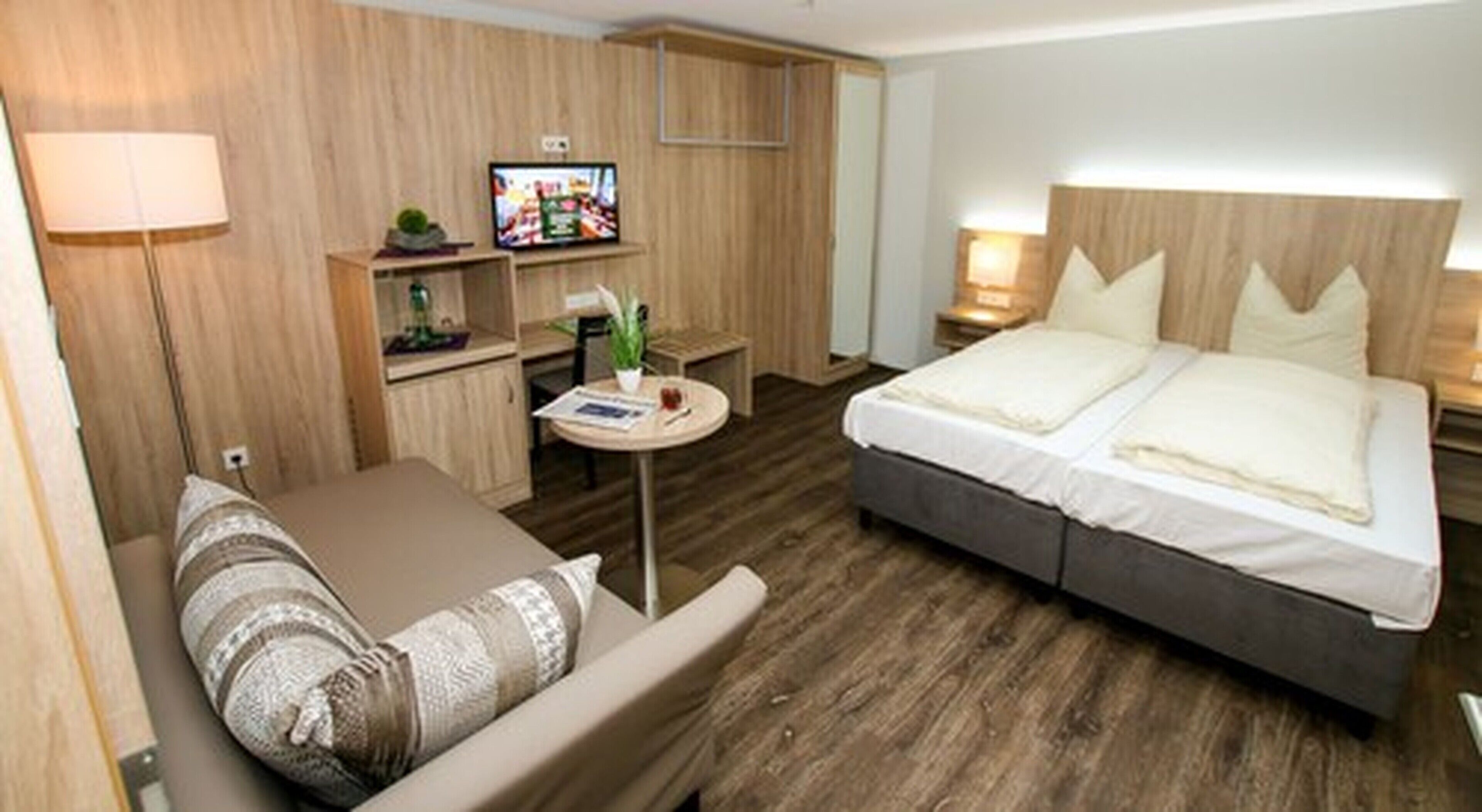 Quarto de hotel compacto com cama, sofá, pequena mesa, televisão e guarda-roupa.
