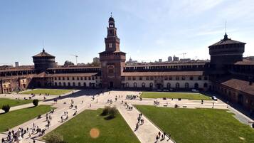 Um pátio com uma torre central, rodeado por edifícios com janelas em arco e um amplo espaço aberto com pessoas.