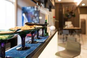 Disposição de buffet com diversos pratos e bebidas, área de bar com copos e um arranjo de assentos com cadeiras.