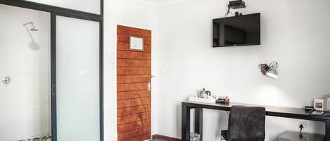 Quarto de hotel com cama, escrivaninha, cadeira, televisão e chuveiro.