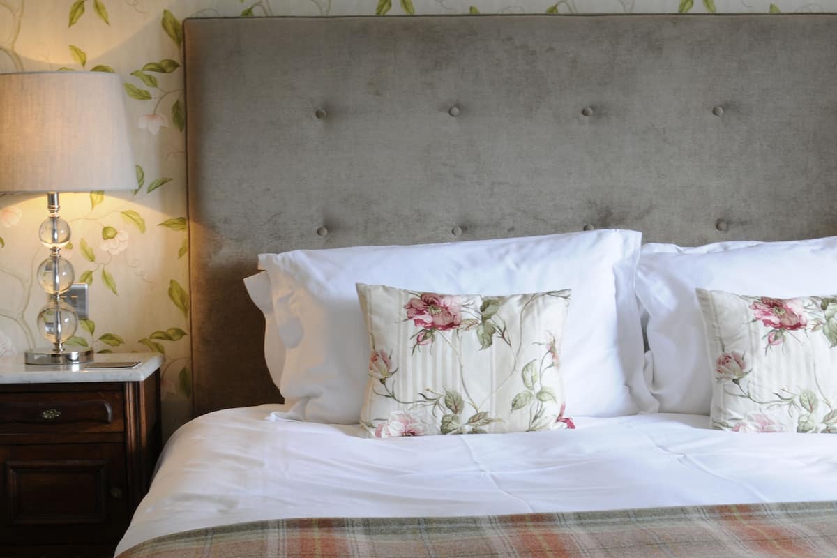 Uma cama bem arrumada com almofadas florais, uma luminária de cabeceira e uma mesa de cabeceira com abajur.
