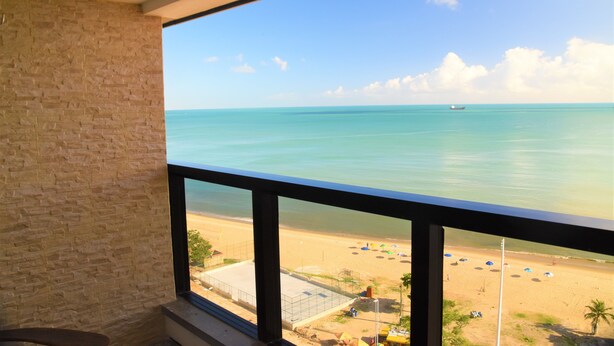 Uma varanda com vista para a praia, céu claro e um barco ao longe.