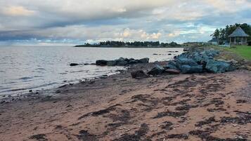 Uma praia com um coreto e cadeiras de descanso, costa rochosa e vista distante de casas e árvores.