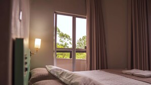 Quarto de hotel com cama, mesa de cabeceira com luminária, janela com cortinas e vista para o verde lá fora.