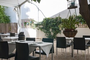 Uma área de refeições ao ar livre com mesas e cadeiras, um vaso suspenso e uma estrutura de tenda branca.
