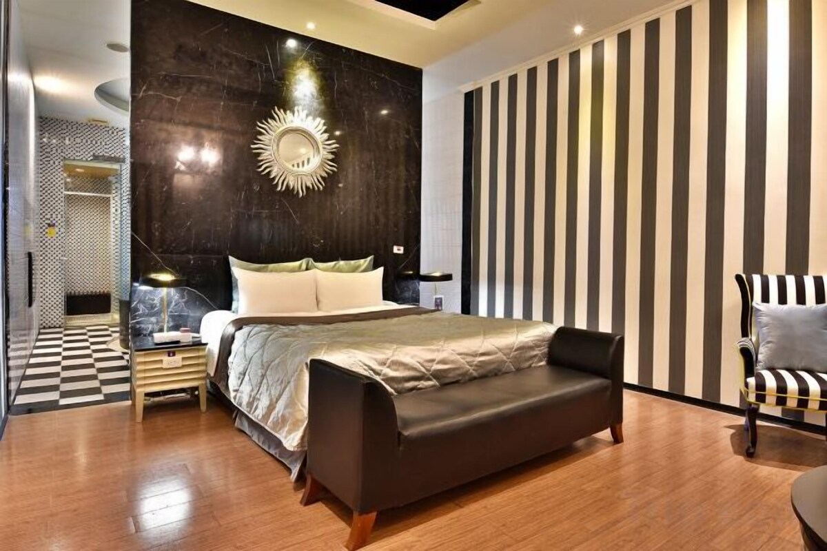 Quarto moderno com uma cama grande, uma mesa de cabeceira, uma cadeira e um luminário fixado na parede.