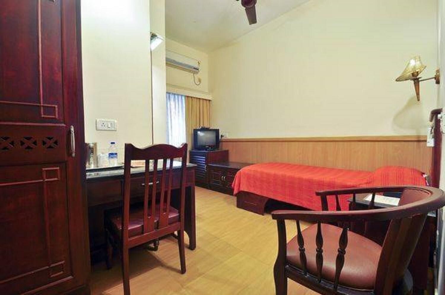 Quarto de hotel com escrivaninha de madeira, uma cadeira, cama com colcha vermelha, televisão e abajur.