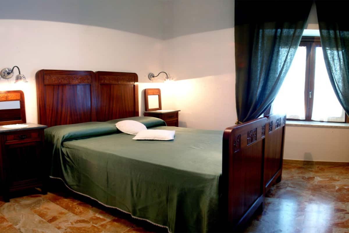 Quarto com cama de madeira, duas mesas de cabeceira com luminárias, um criado-mudo e uma janela com cortinas.