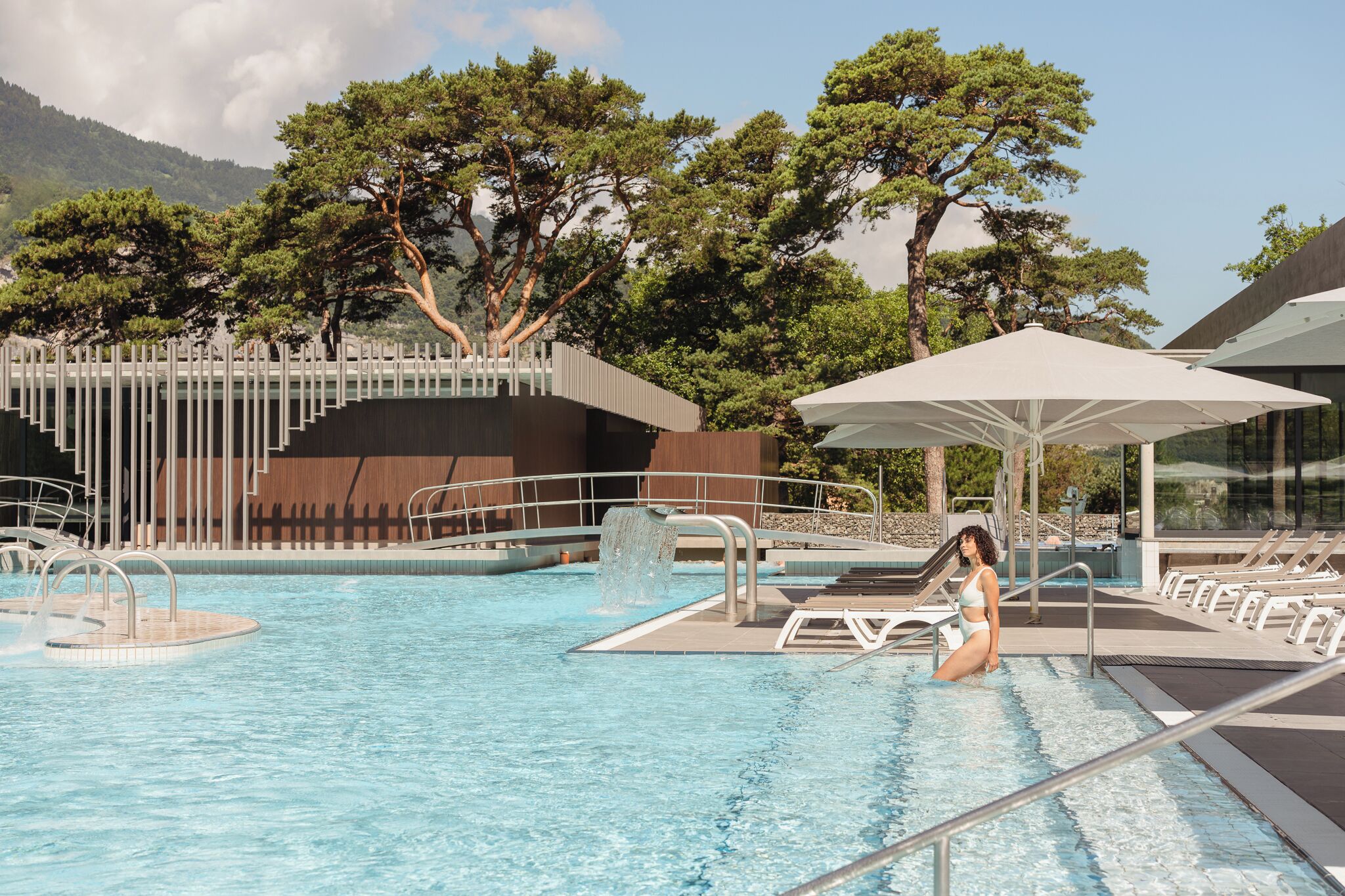Piscina com cachoeira, cercada por árvores e espreguiçadeiras.