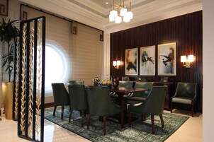 Uma área de refeições com uma mesa redonda, cadeiras verdes, um tapete estampado, obras de arte penduradas na parede e um painel decorativo.